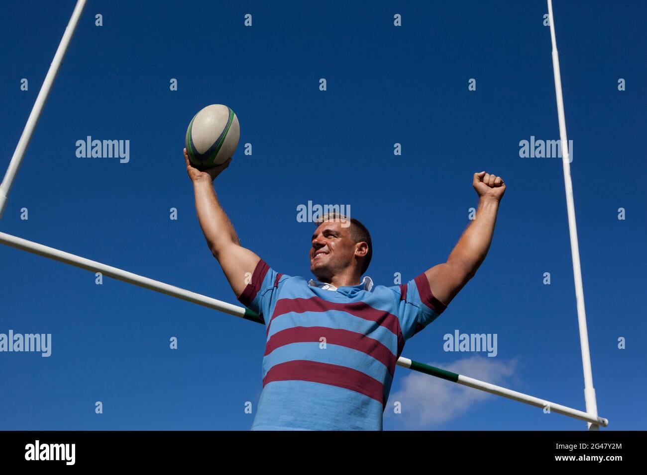 Happy rugby player holding ball with arms raised against blue sky Stock ...