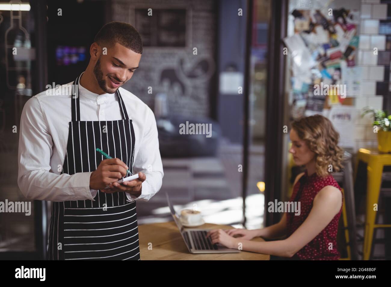 Smiling young waiter writing in notepad while standing at coffee shop ...