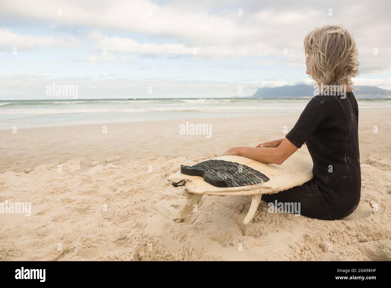 Woman in wetsuit sitting with surfboard against cloudy sky Stock Photo ...