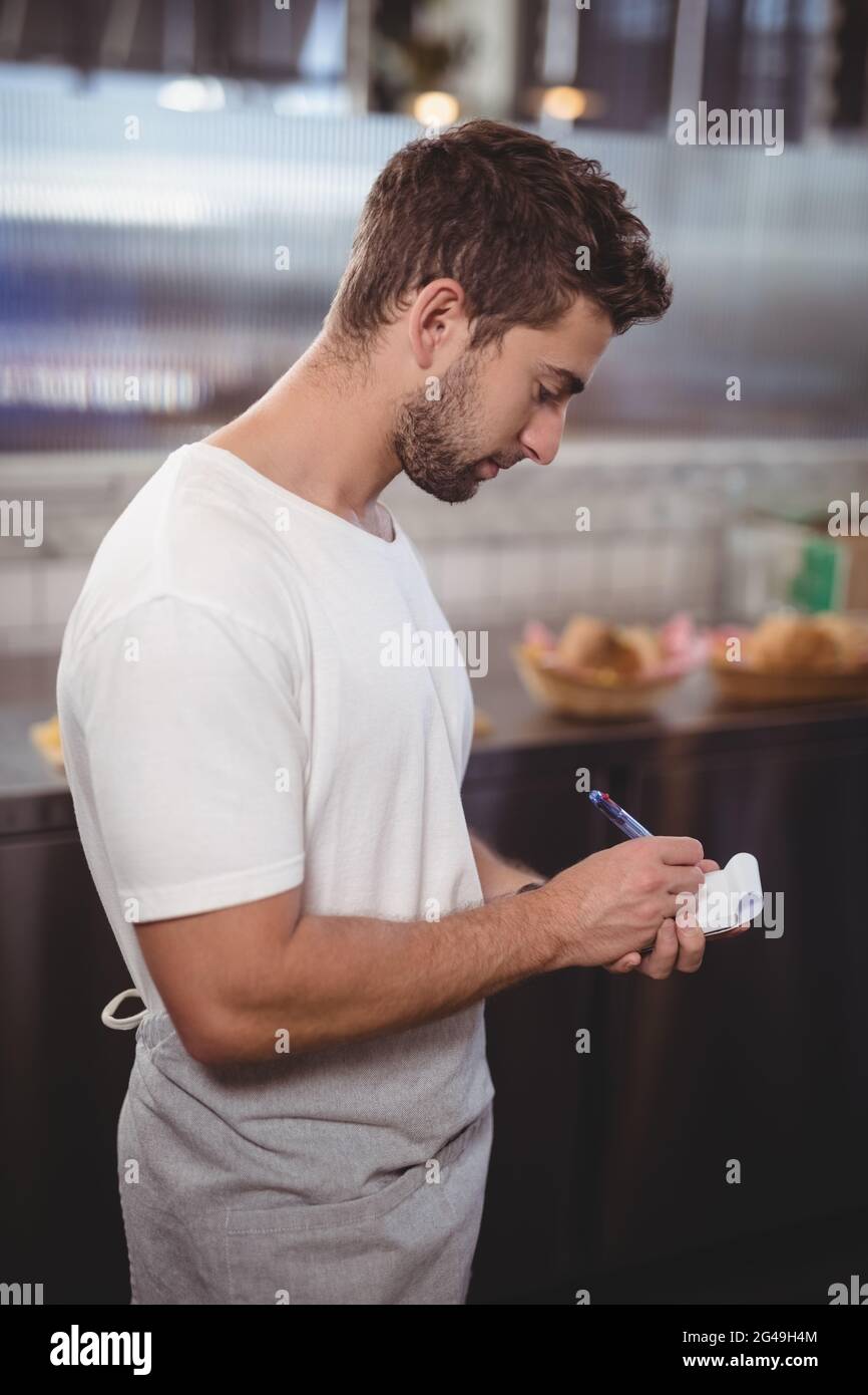 Side view of young handsome waiter writing in notepad at coffee shop ...