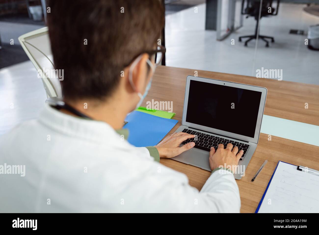 Asian male doctor wearing face mask sitting at desk in hospital office ...