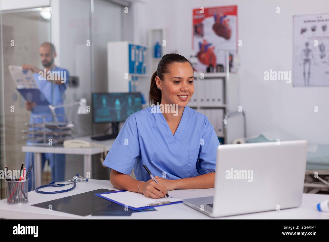 Smiling nurse using laptop computer and writing notes on clipboard in ...
