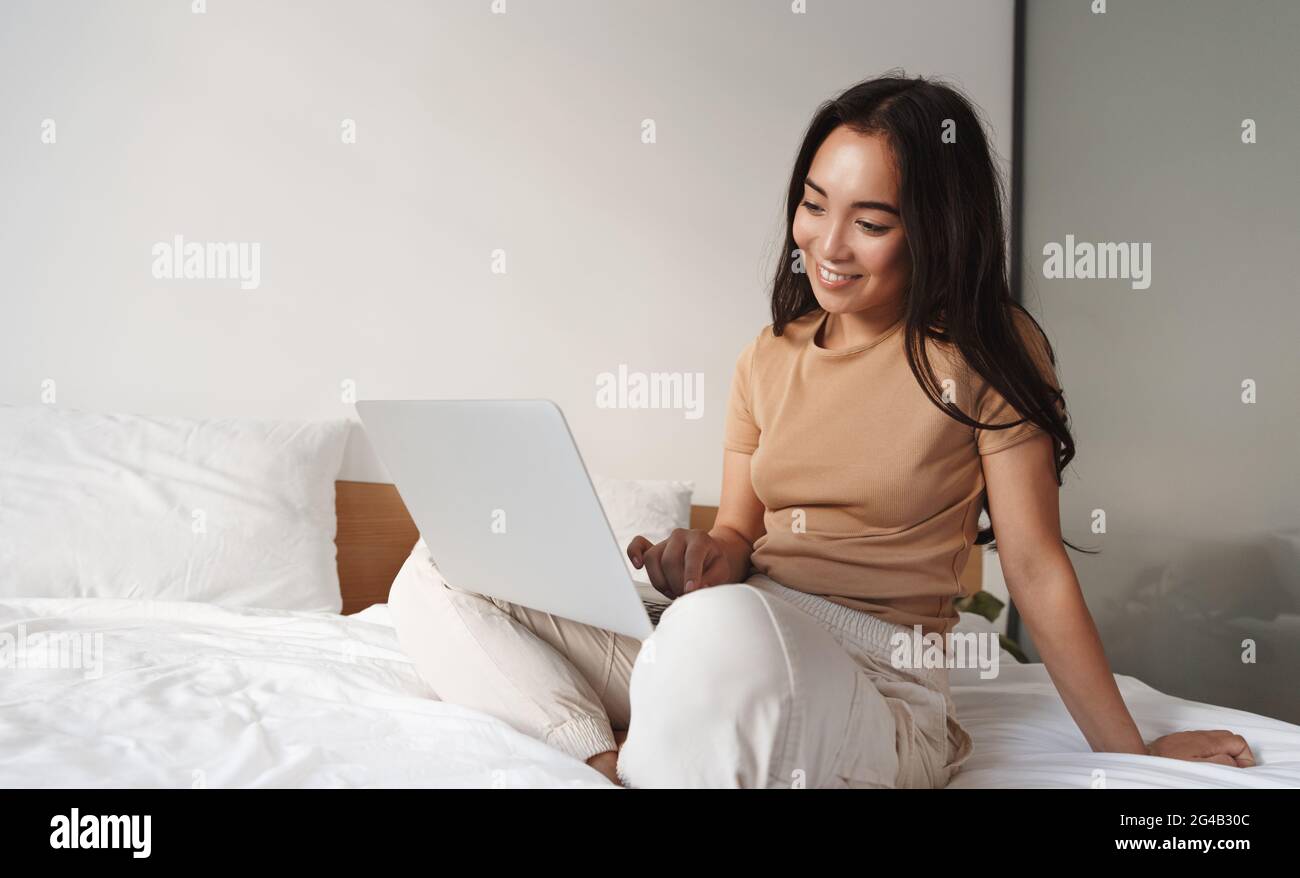 Image of young asian woman using computer in bed. Smiling girl watching ...