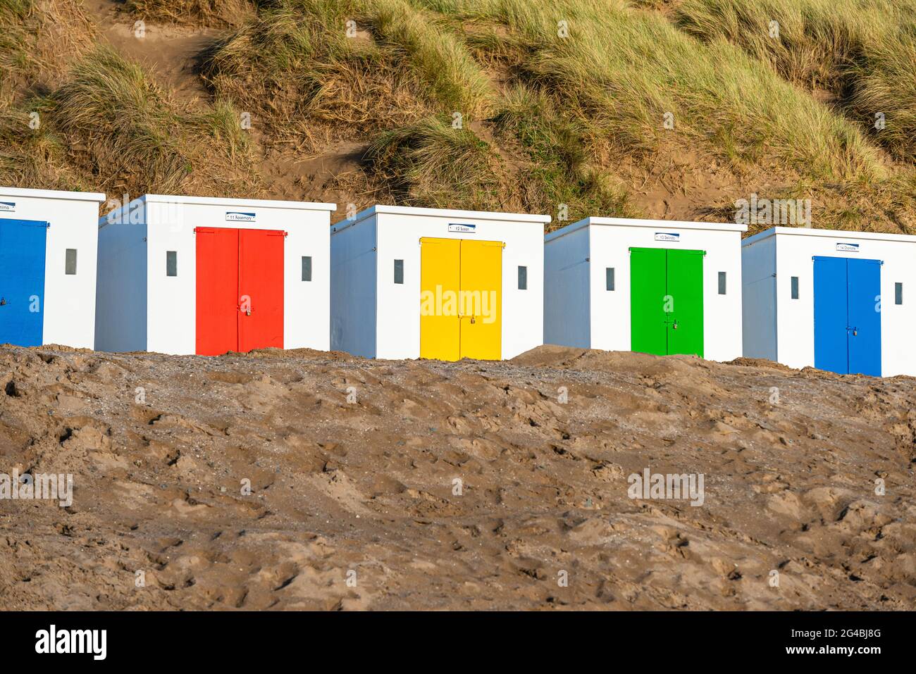 White beach huts, with coloured doors, stand in front of dunes with ...