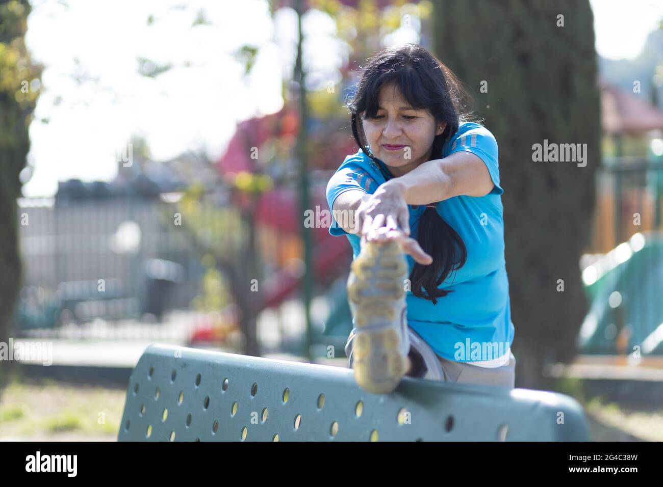 An aged Mexican female in a sportswear is stretching the leg on a park ...