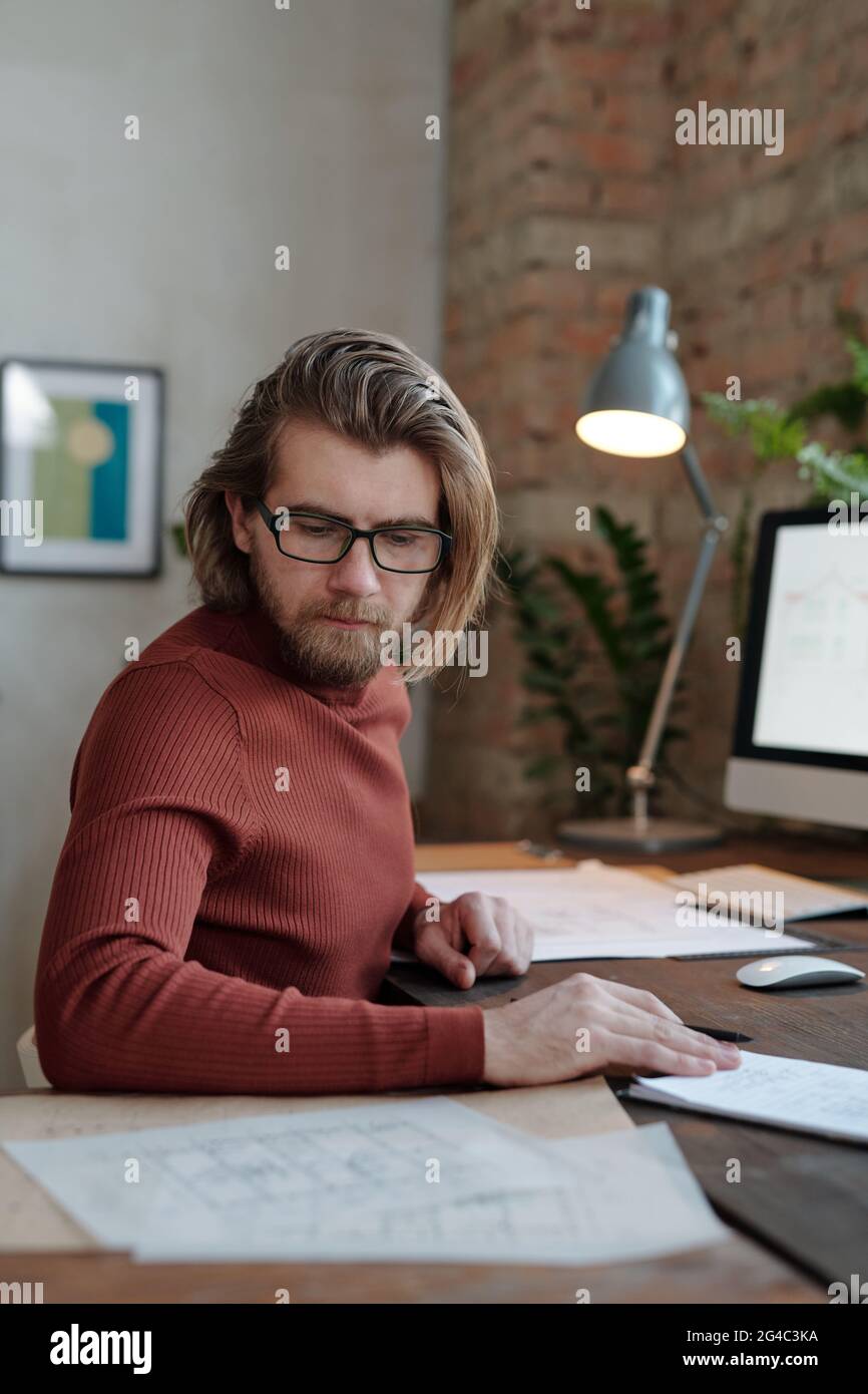Focused young Caucasian bearded architect in glasses sitting at table ...