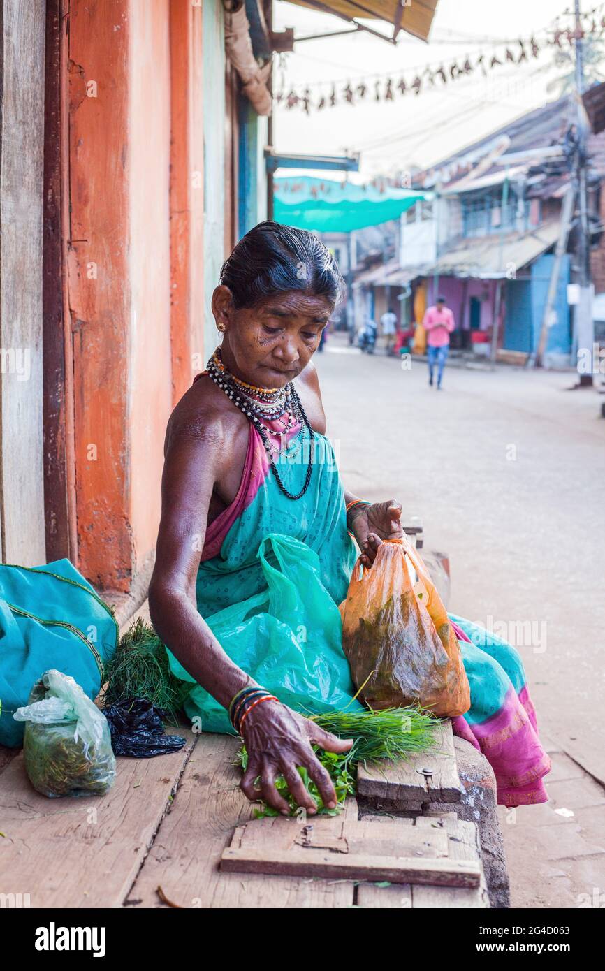 Frail, elderly Indian lady sits on wall on sidewalk arranging green ...
