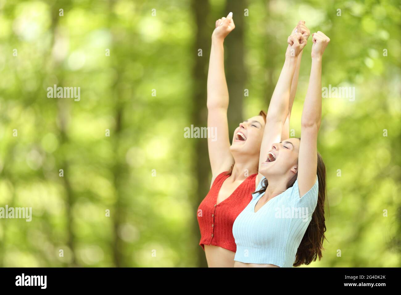 Two excited friends celebrating vacation raising arms in a green forest ...