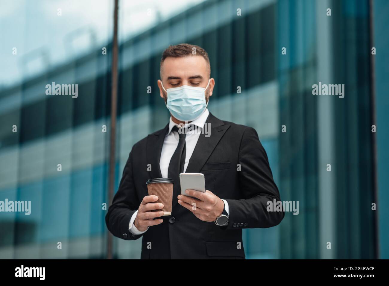 Male entrepreneur in suit wearing face mask and using smartphone, going ...