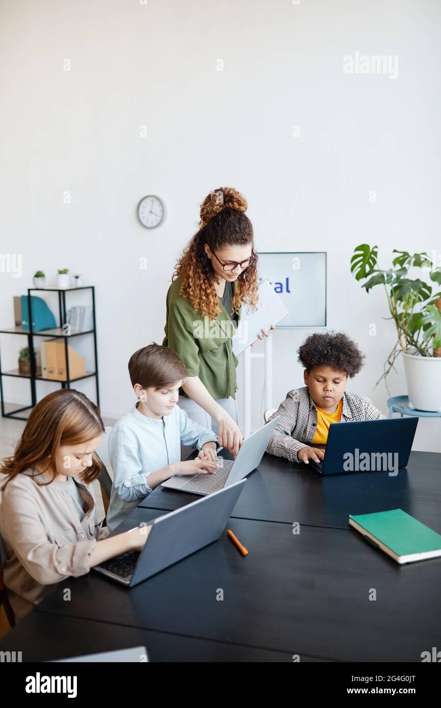 Vertical portrait of young female teacher helping kids using laptops ...