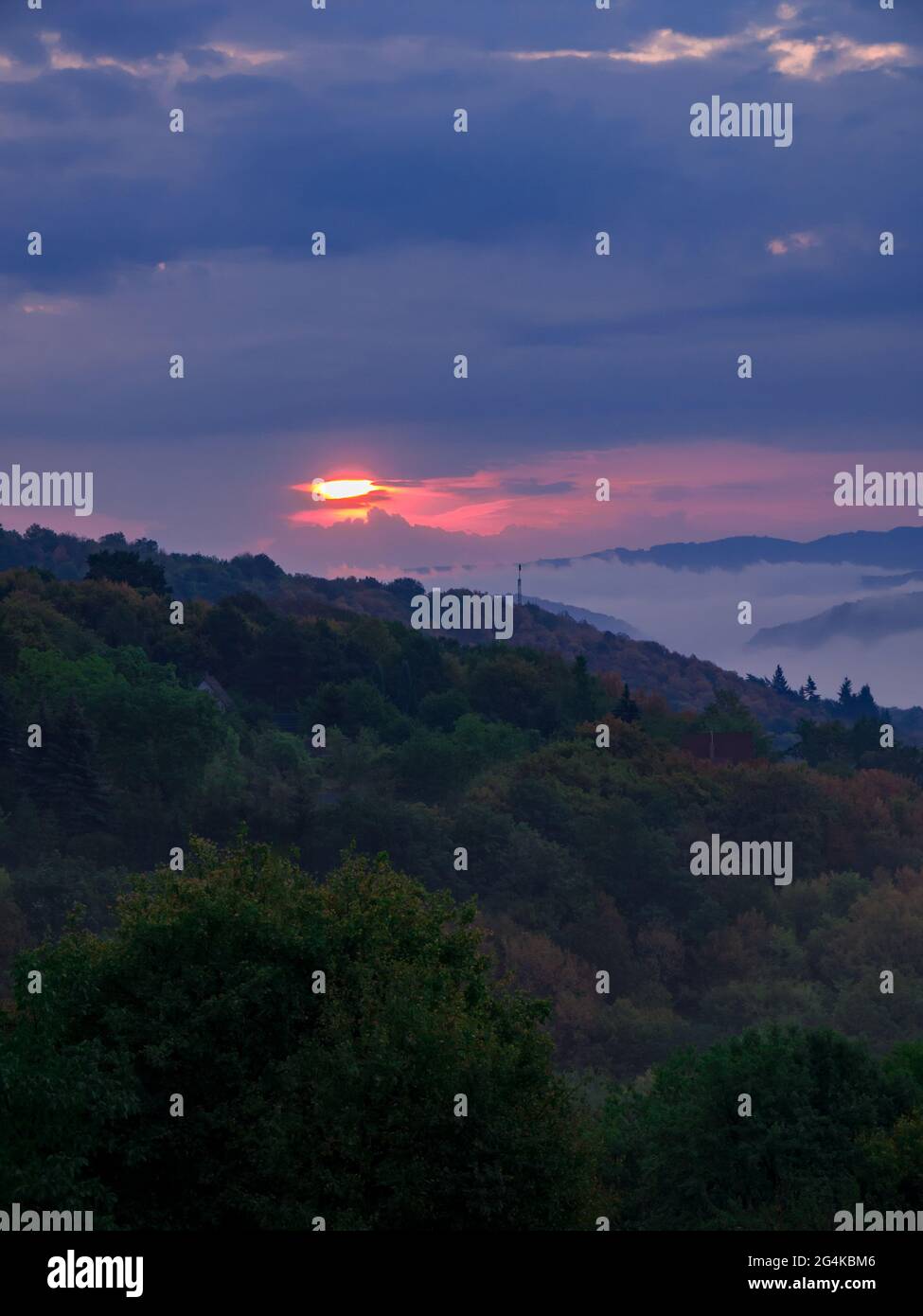 The rising sun peeks from behind heavy rain clouds with a dense fog in ...