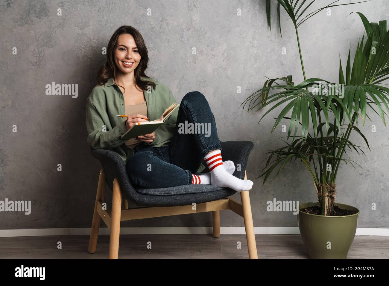 Happy beautiful girl writing down notes while sitting in chair indoors ...