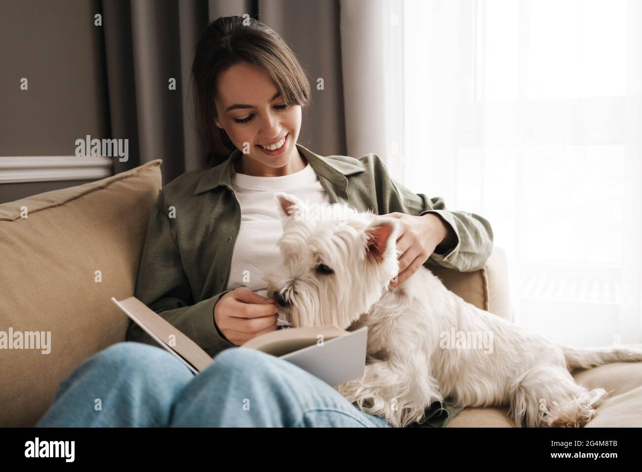 Happy white woman reading book while resting with her dog on sofa at ...