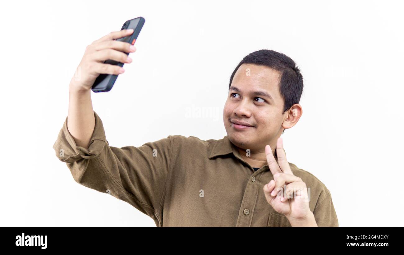 Happy young Asian Malay man taking a selfie and showing a peace sign ...