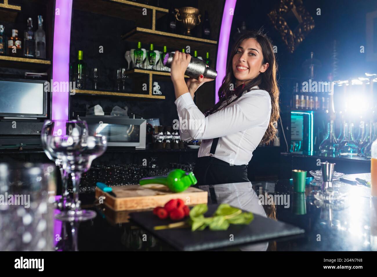 Happy female barkeeper in stylish outfit shaking metal shaker while ...