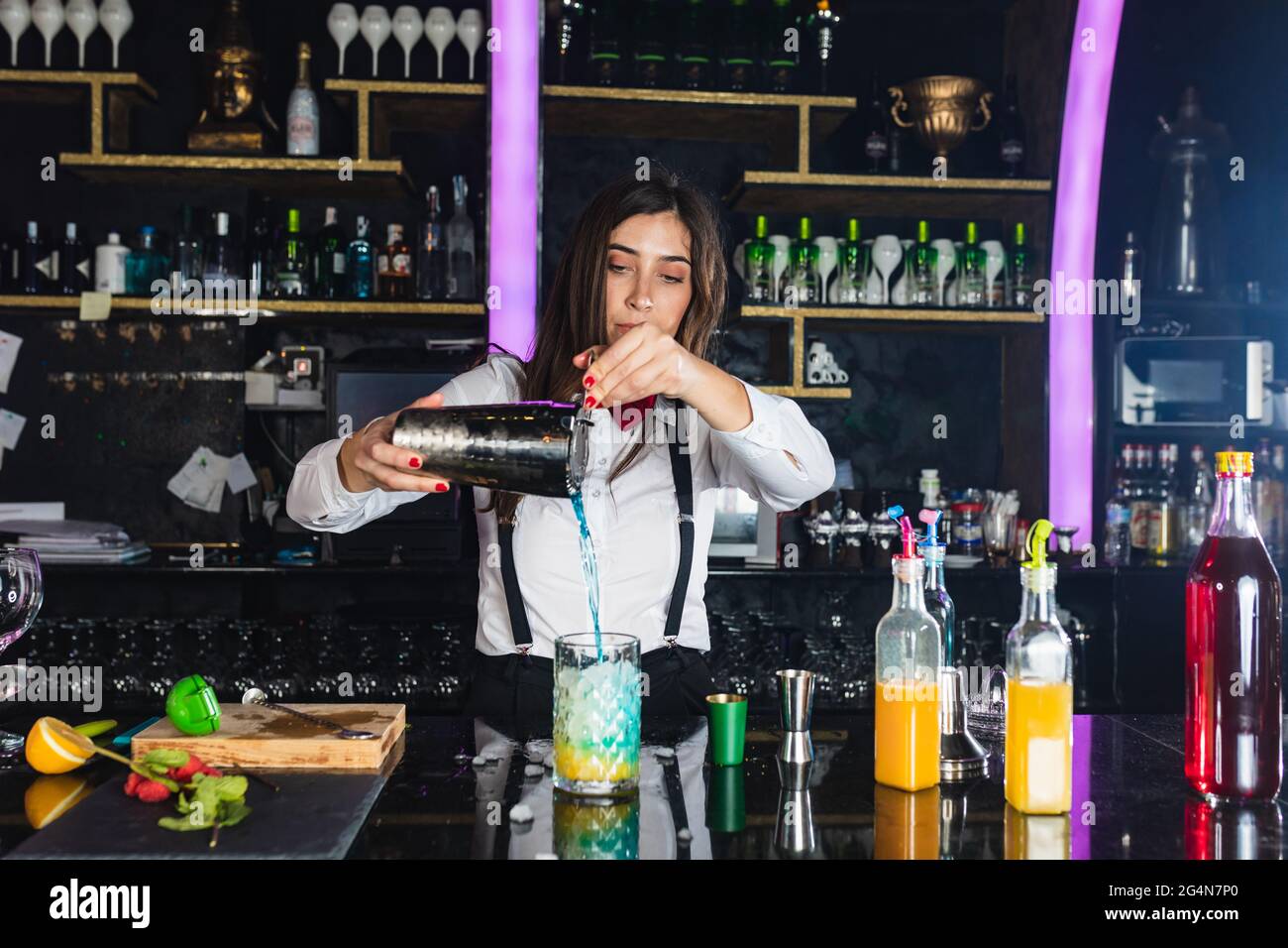 Female barkeeper in stylish outfit serving blue cocktail from a shaker ...