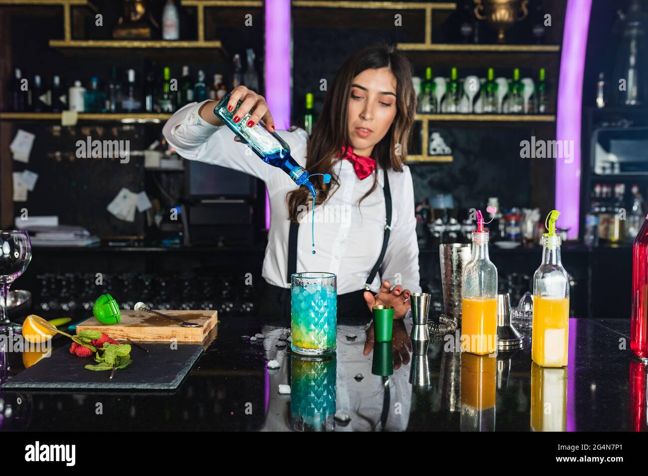 Focused female barkeeper in stylish outfit adding blue colorant liquid ...