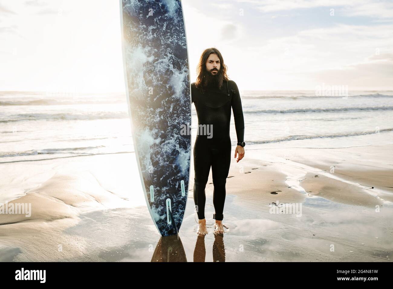 Surfer man with long hair and beard dressed in wetsuit standing looking ...