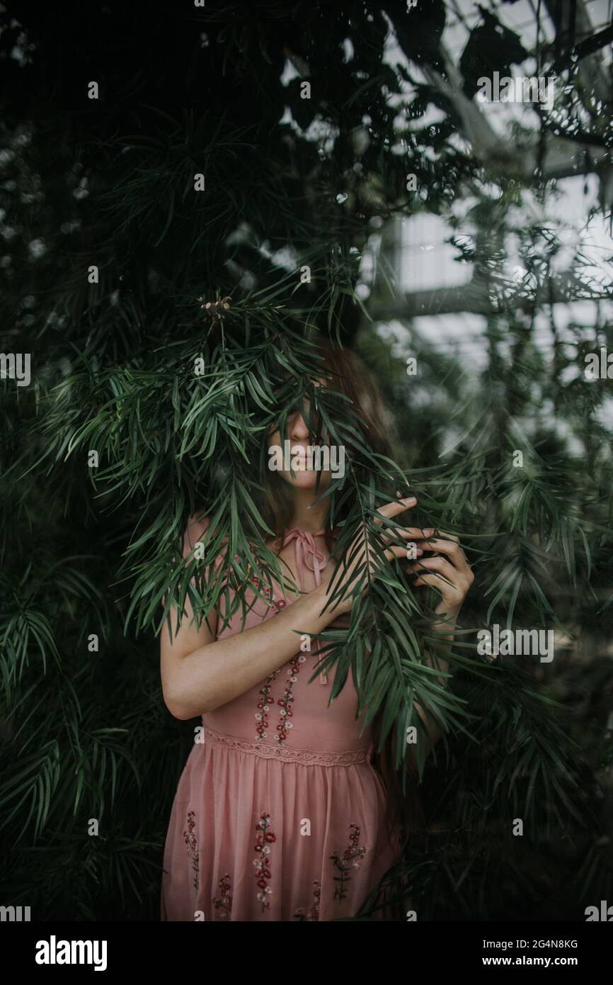 Female standing in lush greenhouse and hiding face behind branch of ...
