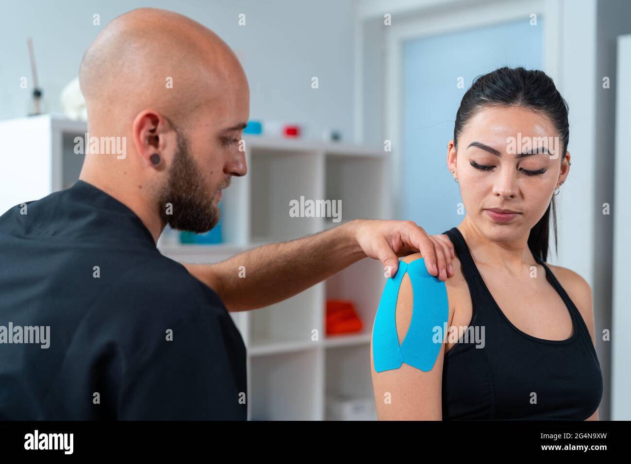 Bearded male physiotherapist applying kinesio tapes on shoulder of female athlete during ...
