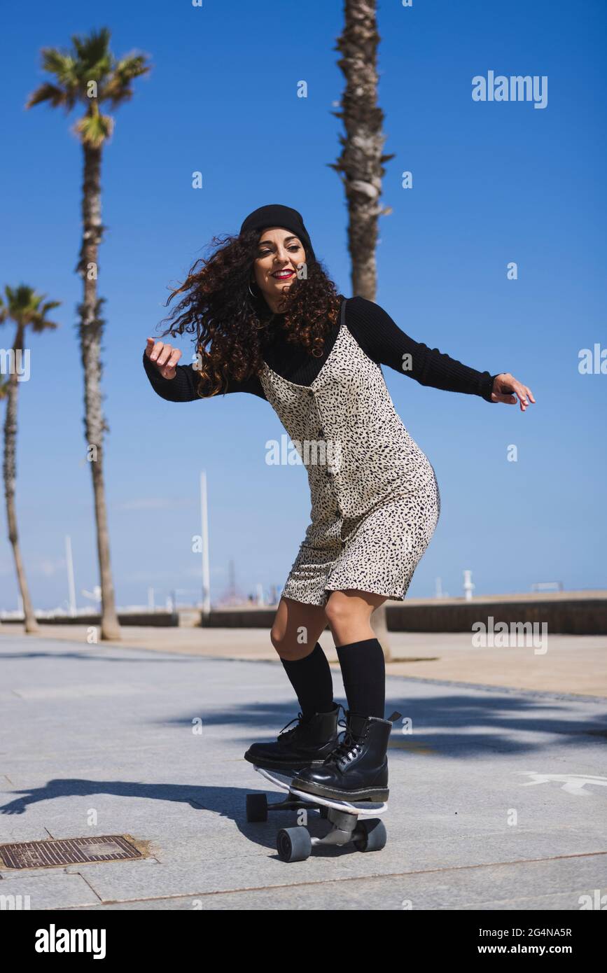 Full body of active happy female in dress riding skateboard on road ...