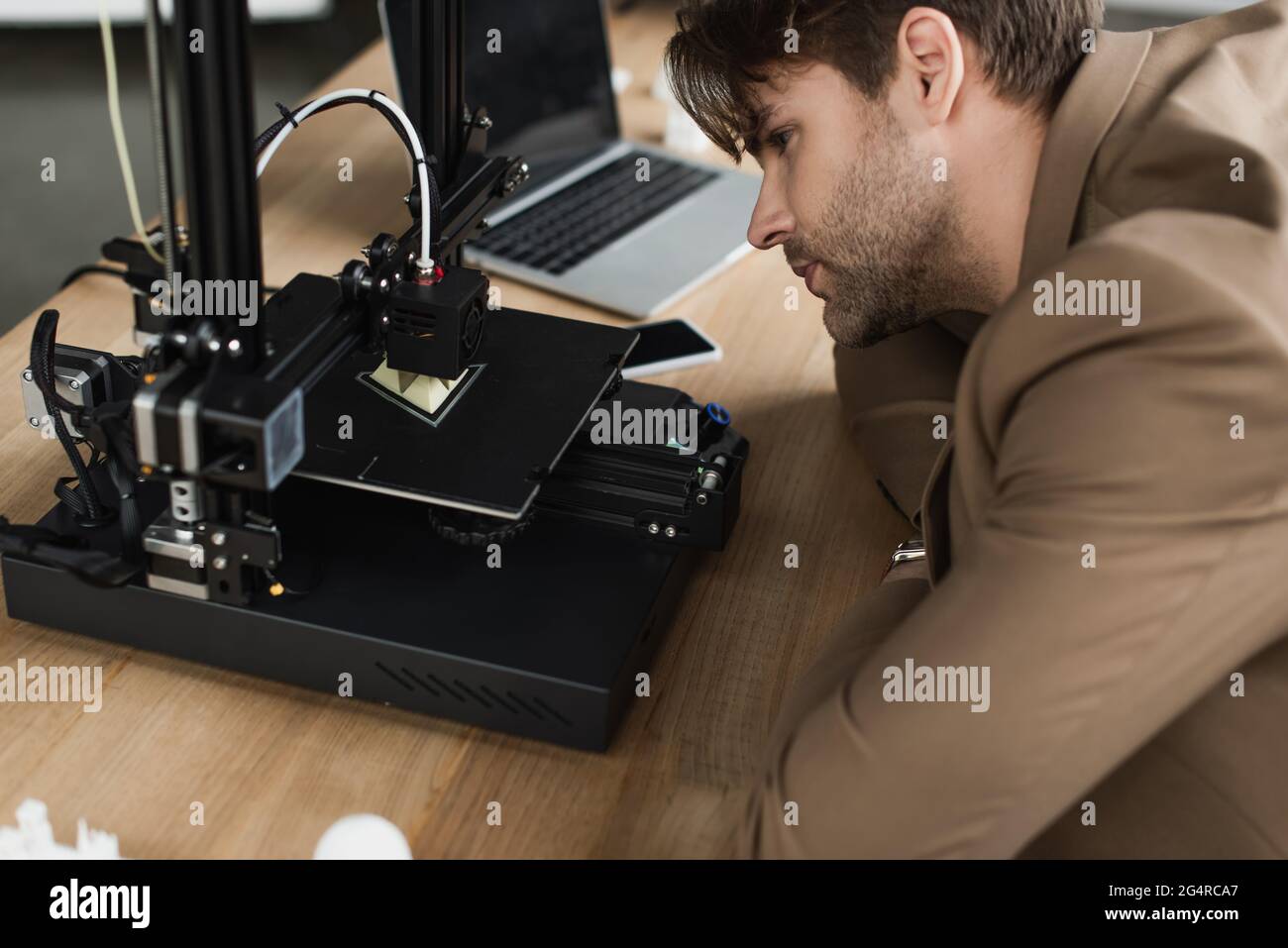 concentrated young man looking at 3D printer with plastic figure in ...