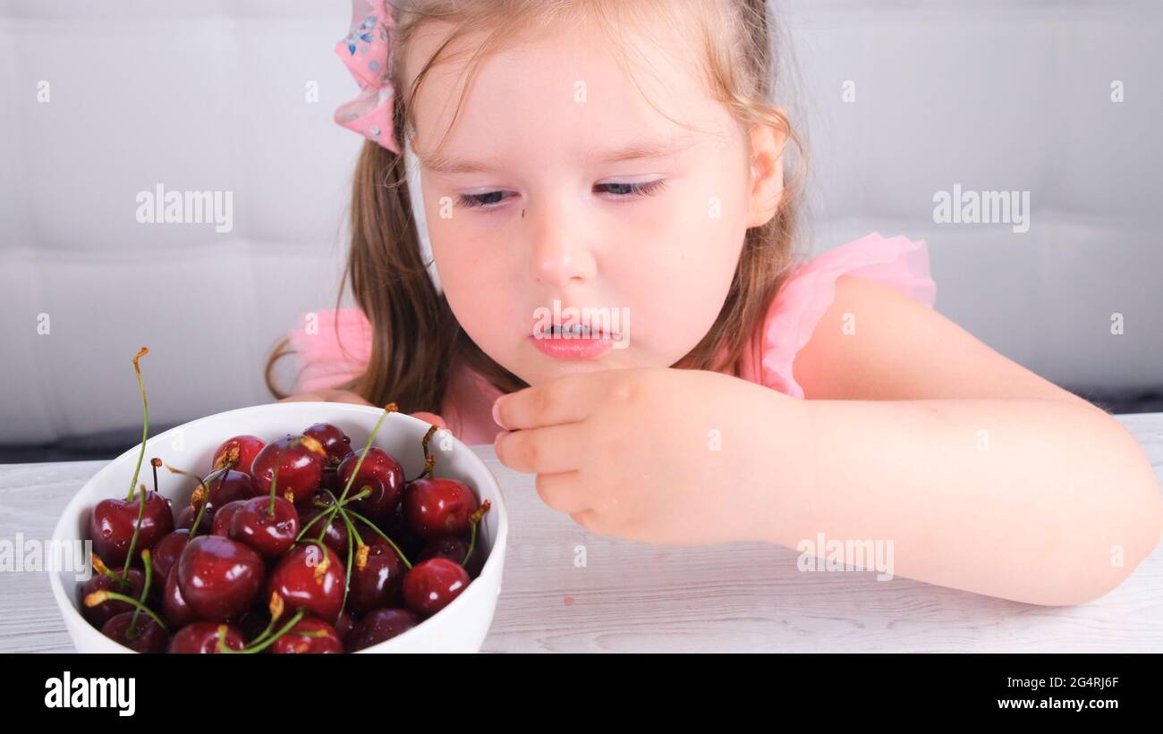 Beautiful smiling little girl eating cherries over white. little cute girl sitting with a plate ...