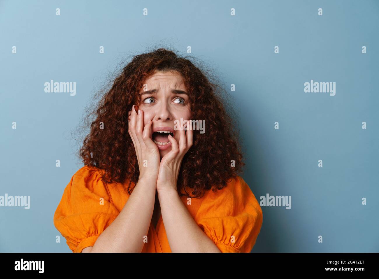 Scared ginger woman screaming and touching her face isolated over blue ...