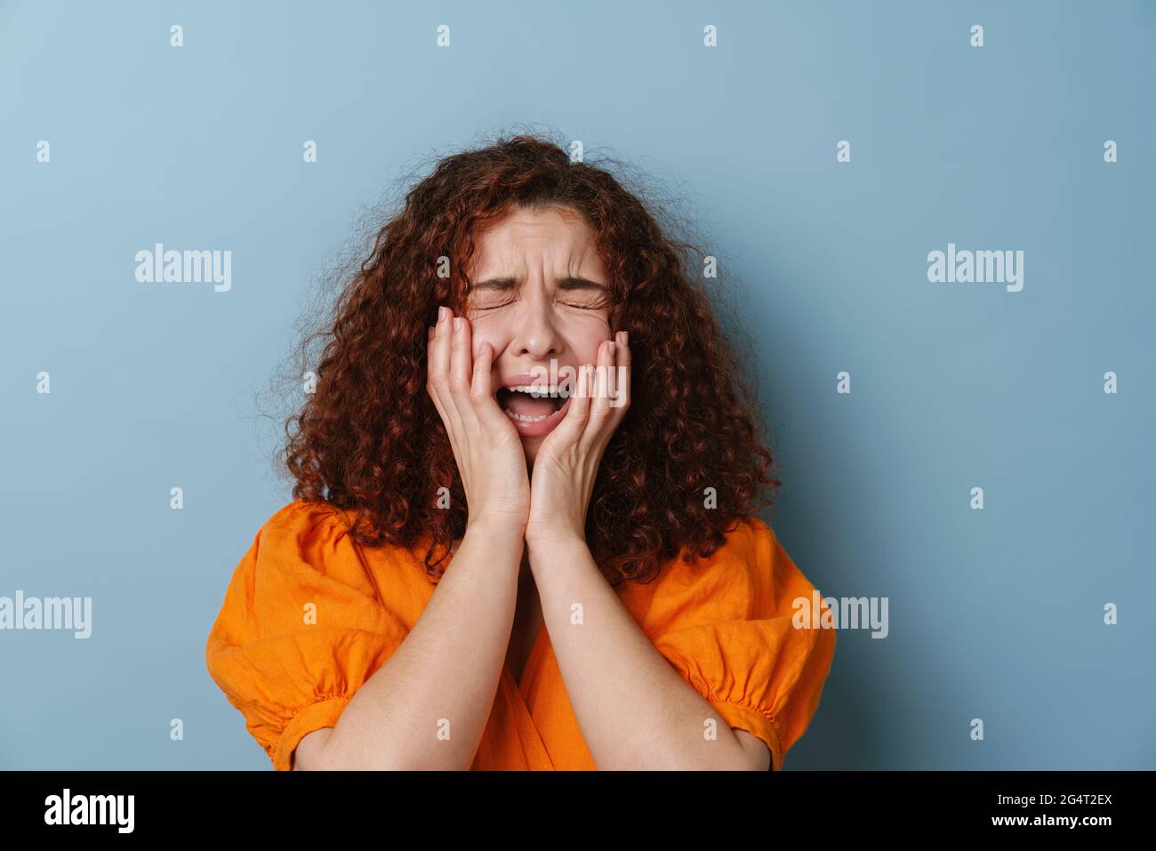 Scared ginger woman screaming and touching her face isolated over blue ...