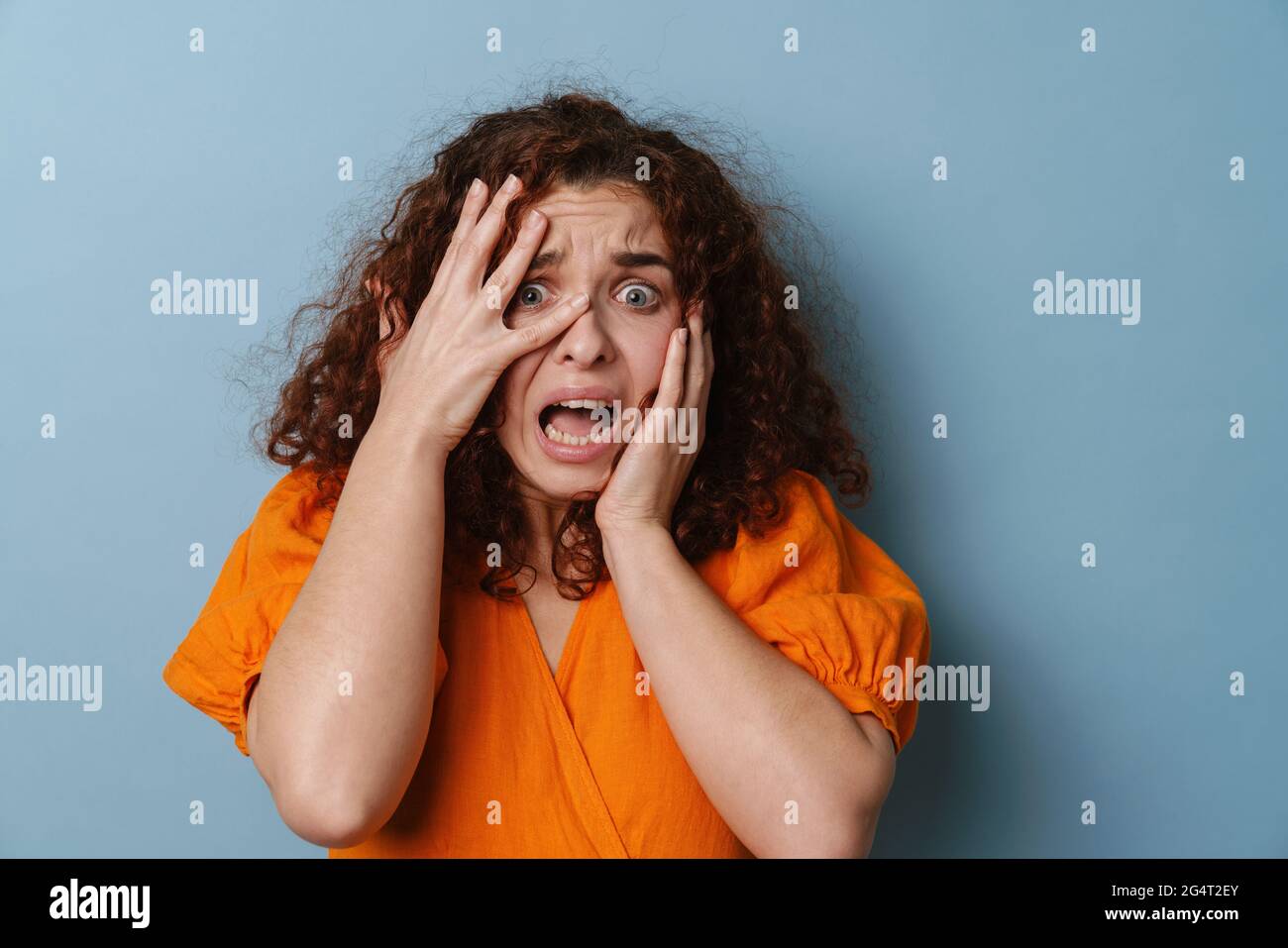 Scared ginger woman grimacing and covering her face isolated over blue ...