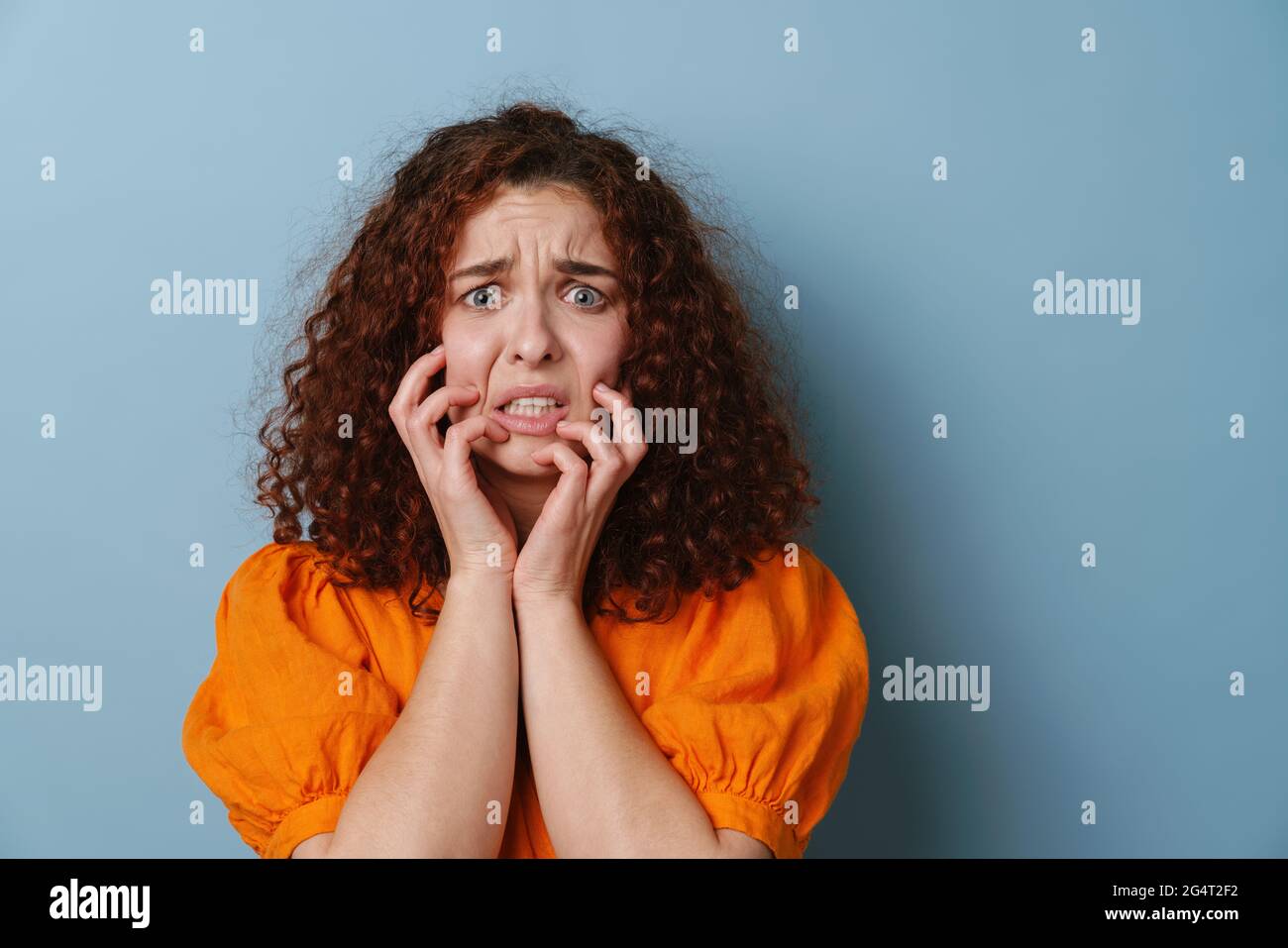 Scared ginger woman grimacing and touching her face isolated over blue ...