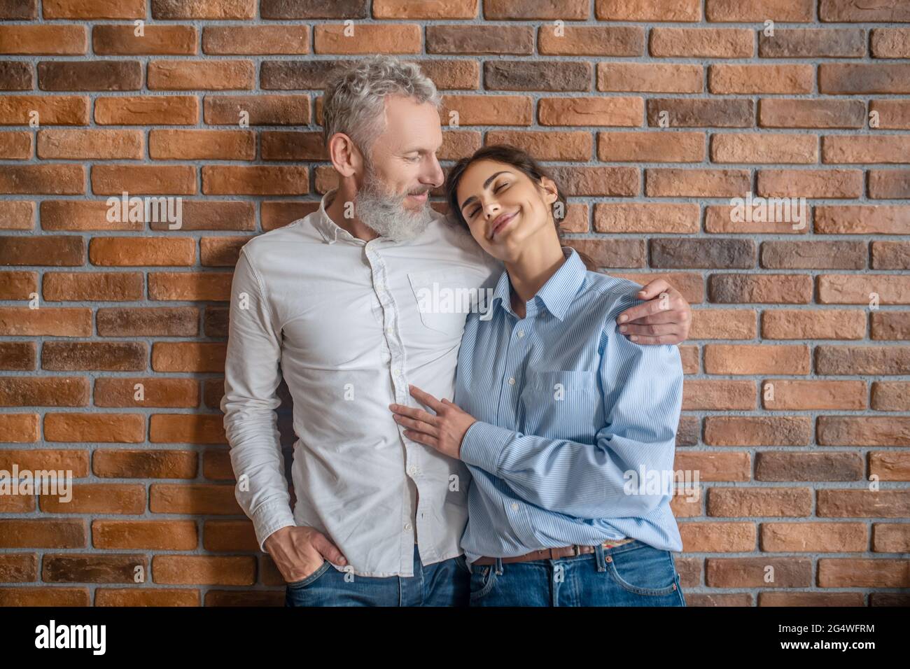 A Gray Haired Man Hugging His Wife While Standing Near The Brick Wall