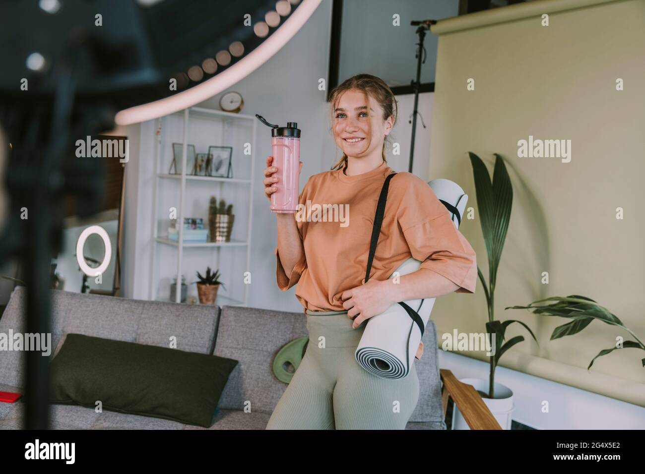 Smiling female influencer with exercise mat and smoothie vlogging at ...