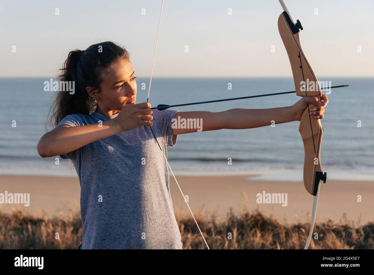 Young woman aiming with bow and arrow while standing at beach Stock ...
