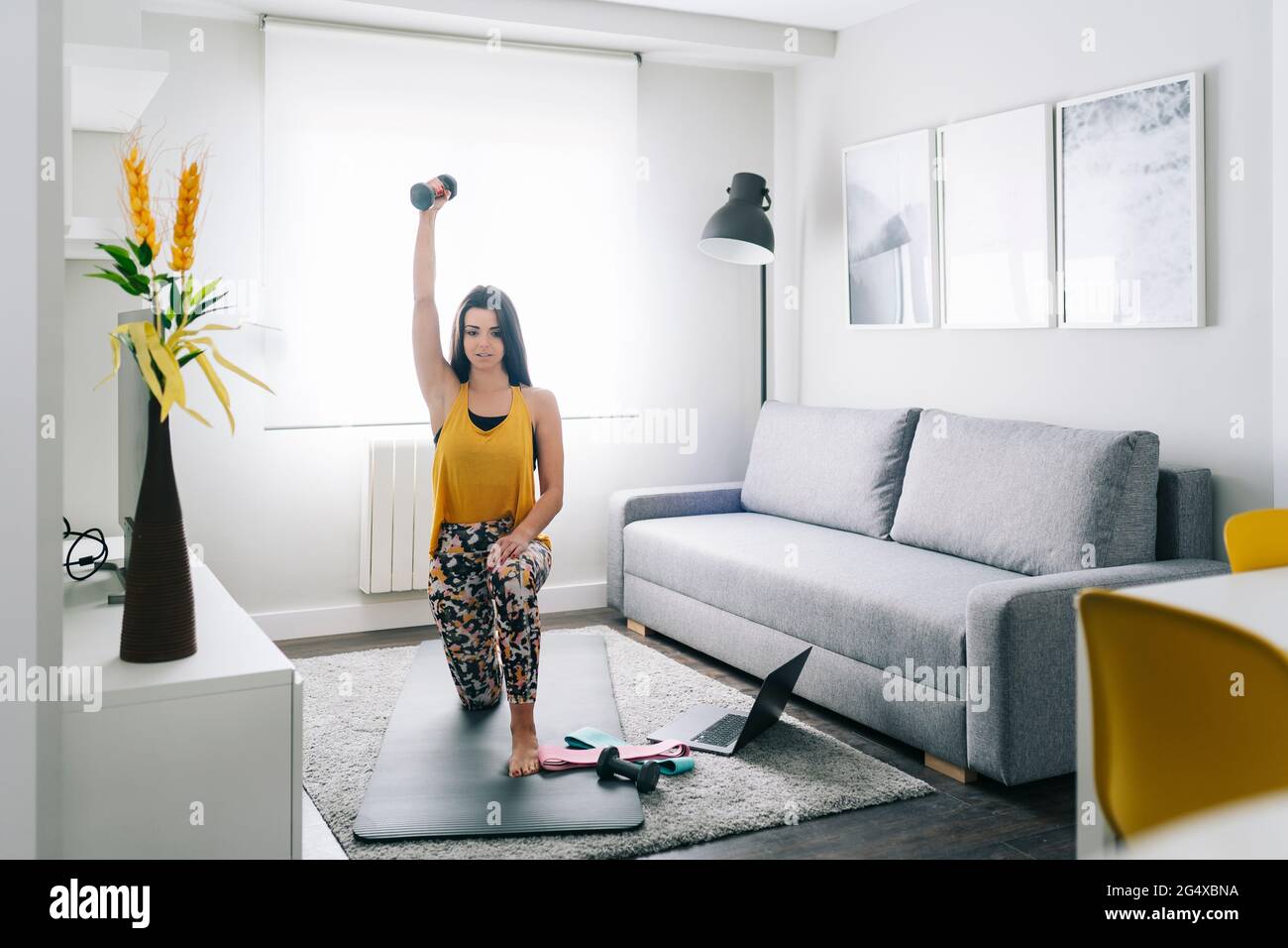 Young woman exercising with dumbbell while crouching on exercise mat at ...