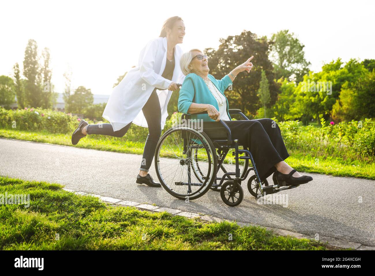 Cheerful senior woman pointing while caretaker wheeling wheelchair in ...