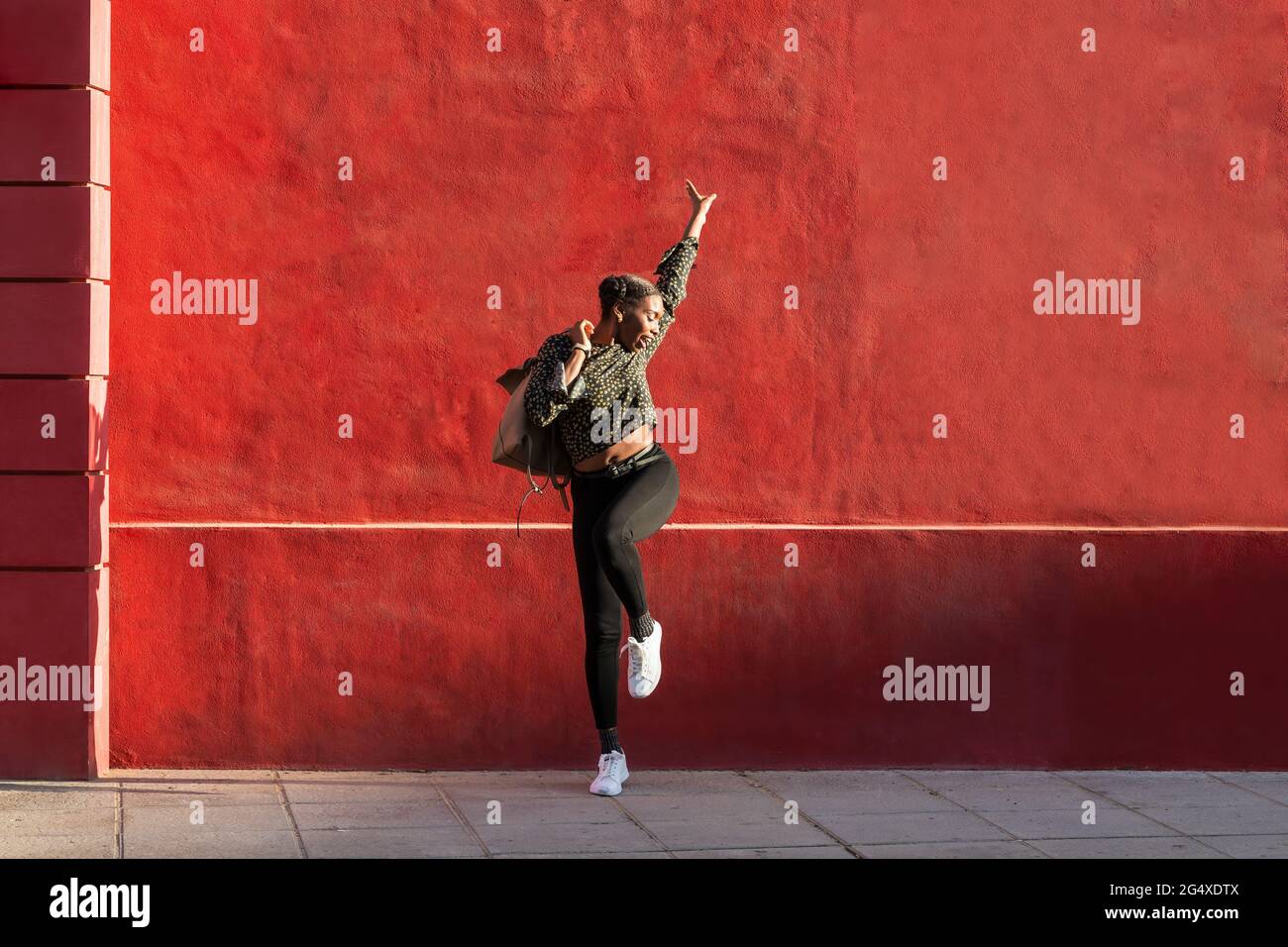 Carefree woman standing with hand raised standing in front of wall ...
