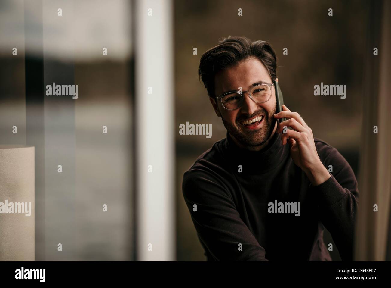 Happy man talking on mobile phone while looking through window Stock ...