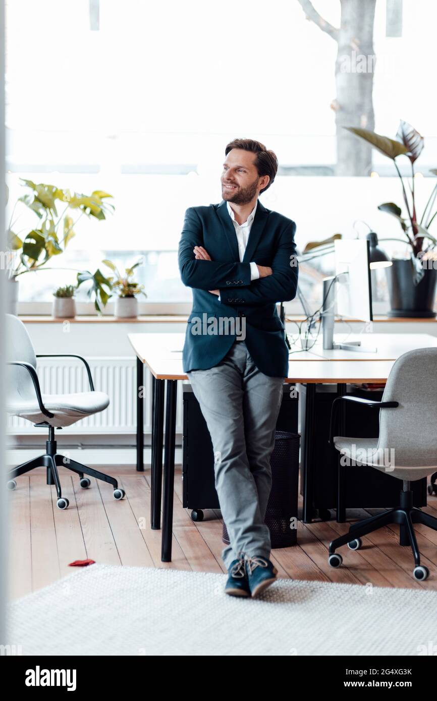 Businessman with arms crossed leaning on desk in office Stock Photo - Alamy