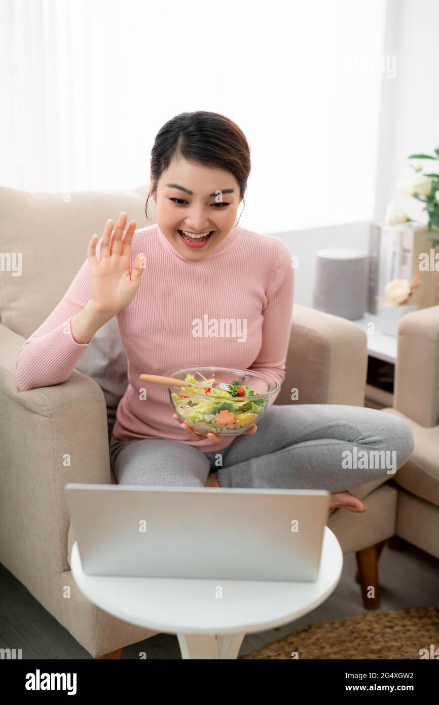 Beautiful young woman sitting on sofa, eating a healthy salad, using a ...