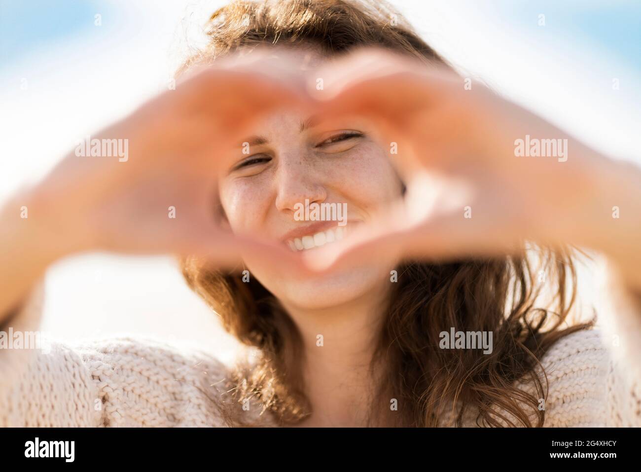 Happy young woman looking through heart shape made by hand on beach ...