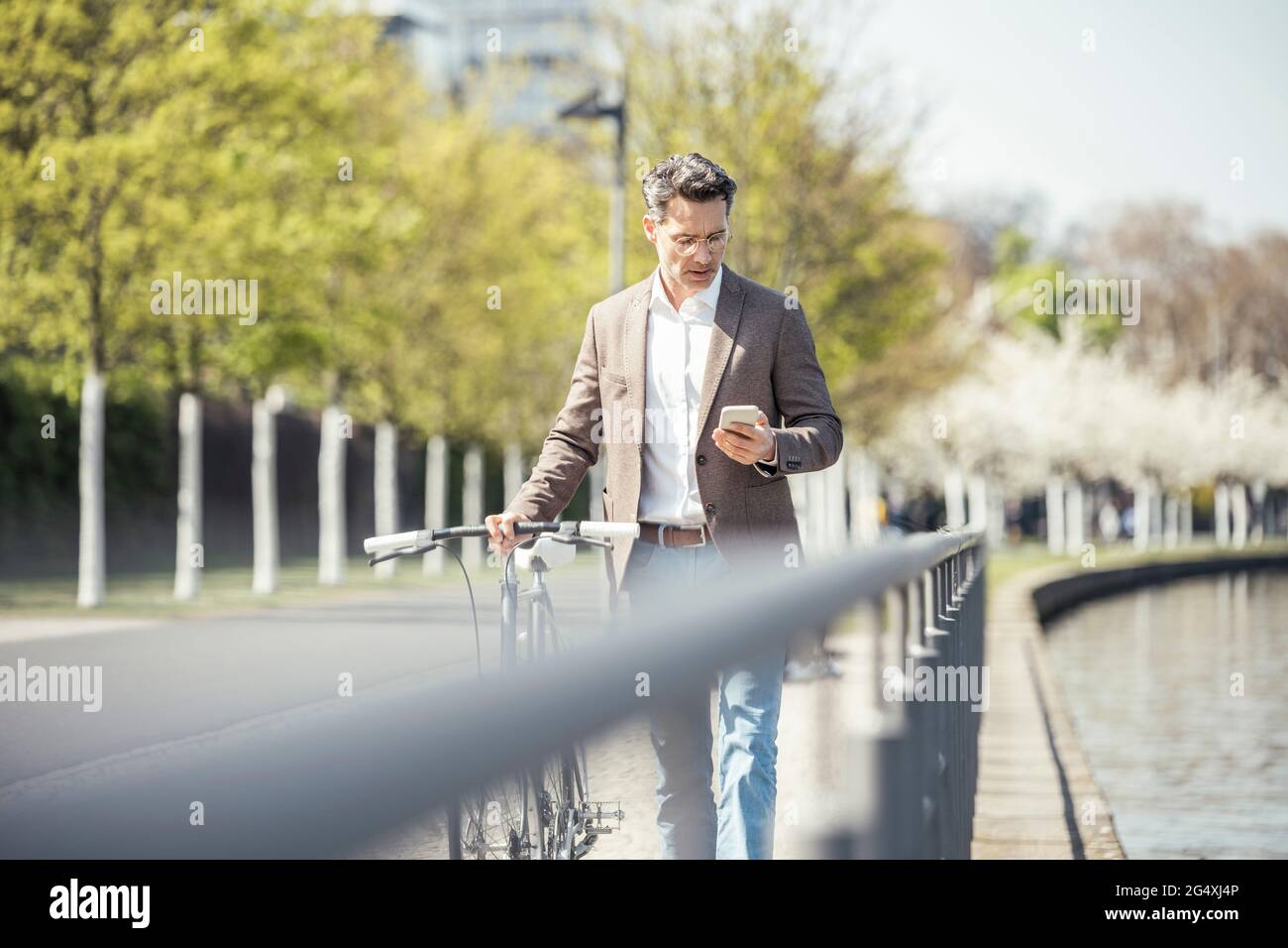 Mature businessman using mobile phone while walking with bicycle by ...