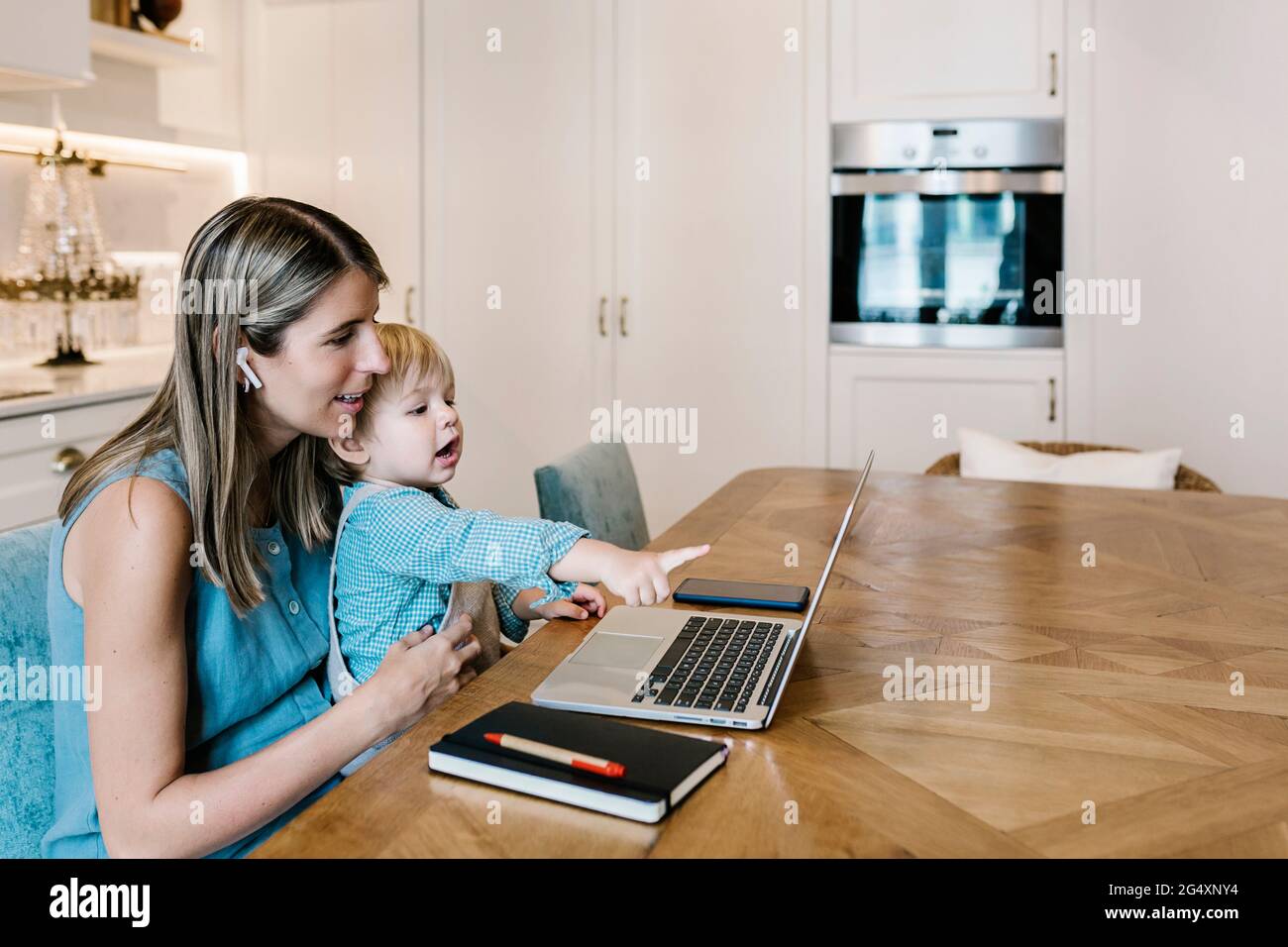 Boy pointing at laptop while sitting on mother's lap working in kitchen ...