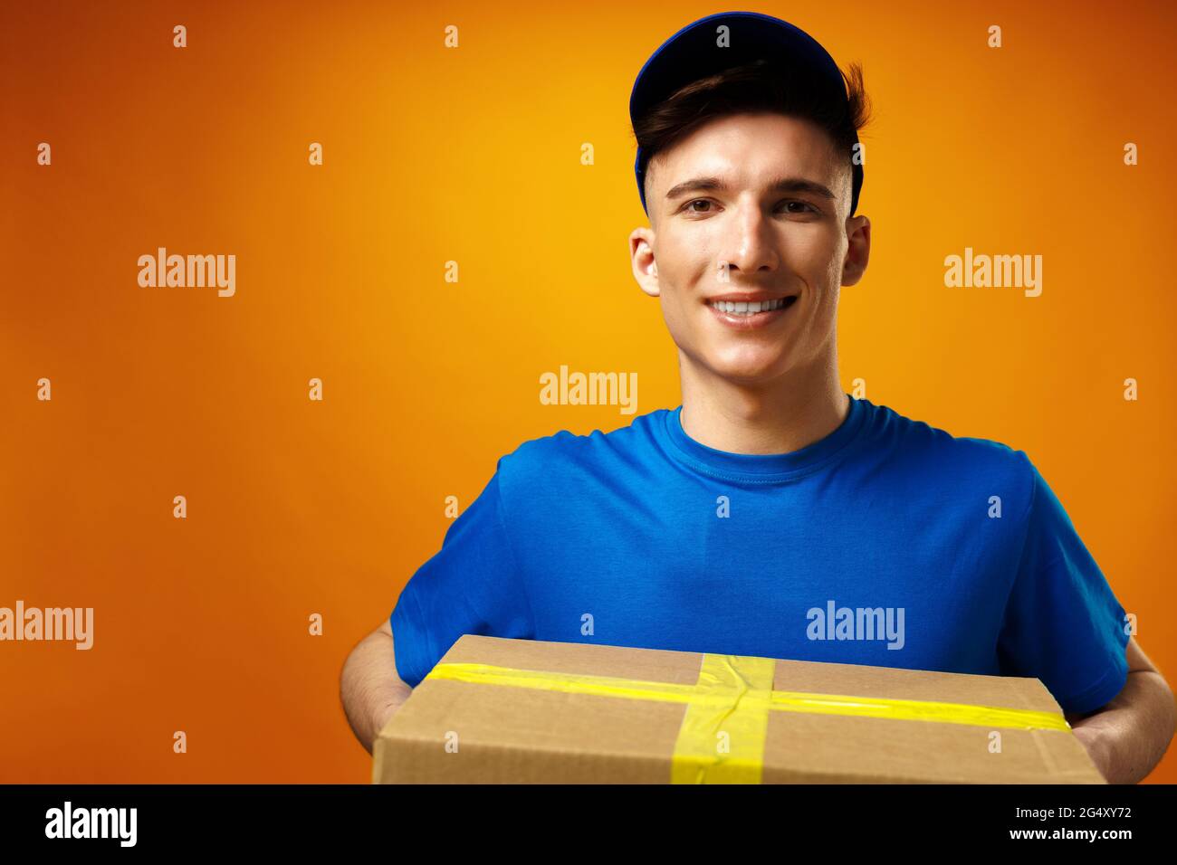 Young handsome delivery man holding parcel cardboard box against yellow ...