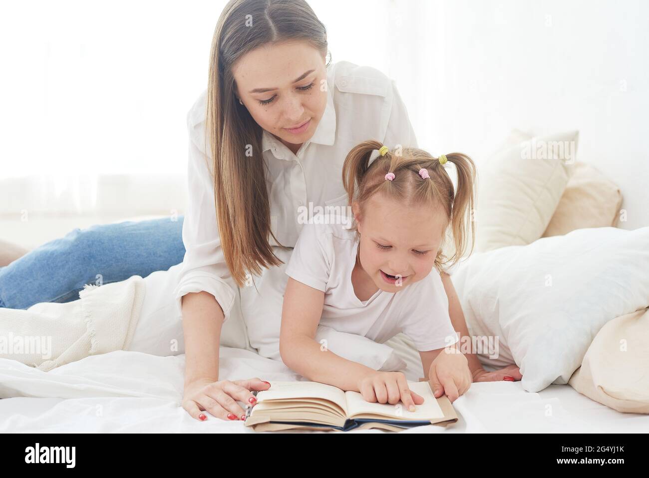 Smiling young mother or nanny sit in bed with cute little girl child ...