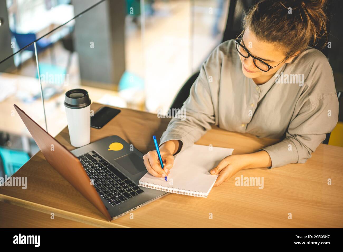 A woman in glasses studying online with her laptop, writing in notepad ...