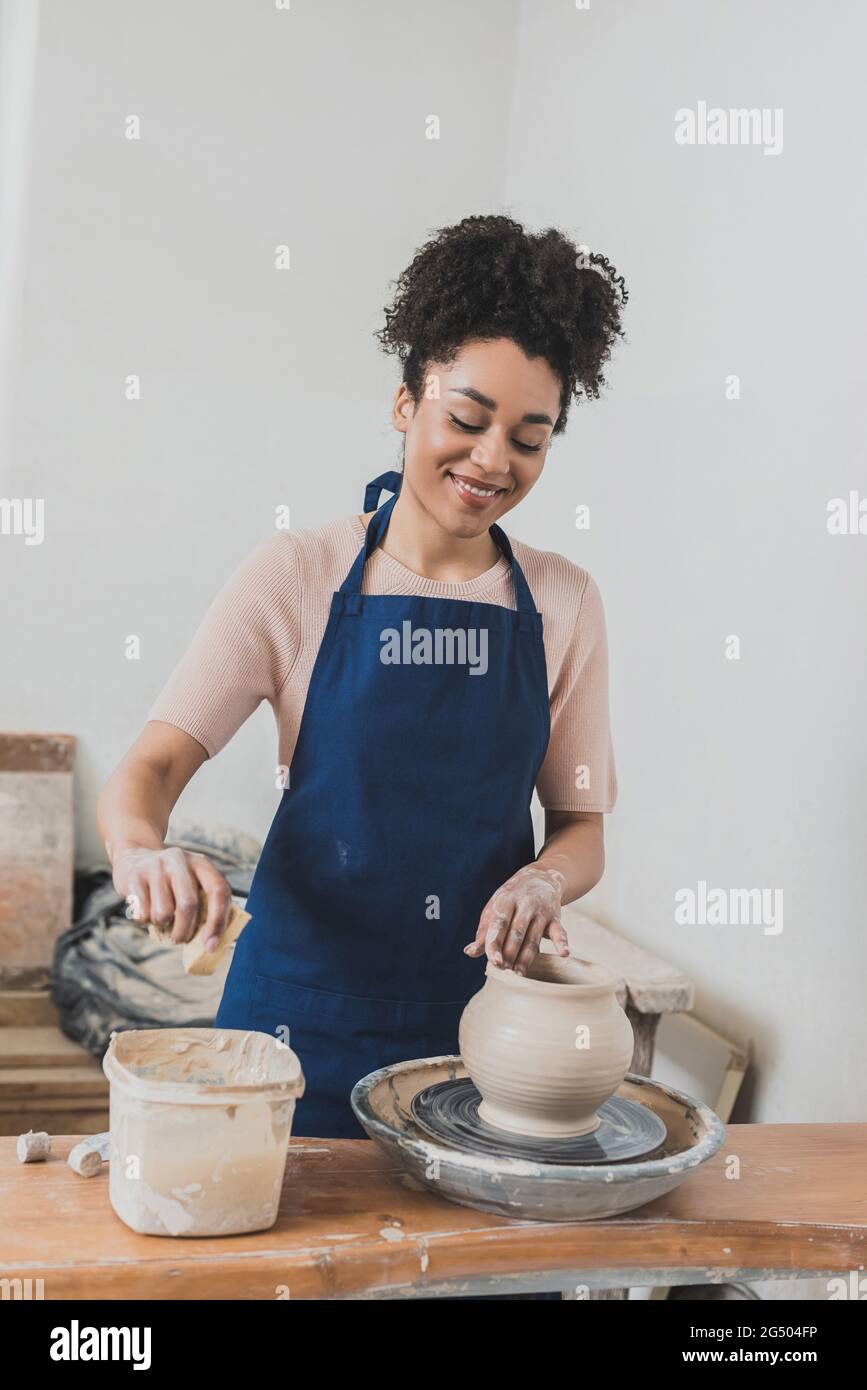 smiling young african american woman in apron modeling wet clay pot on ...