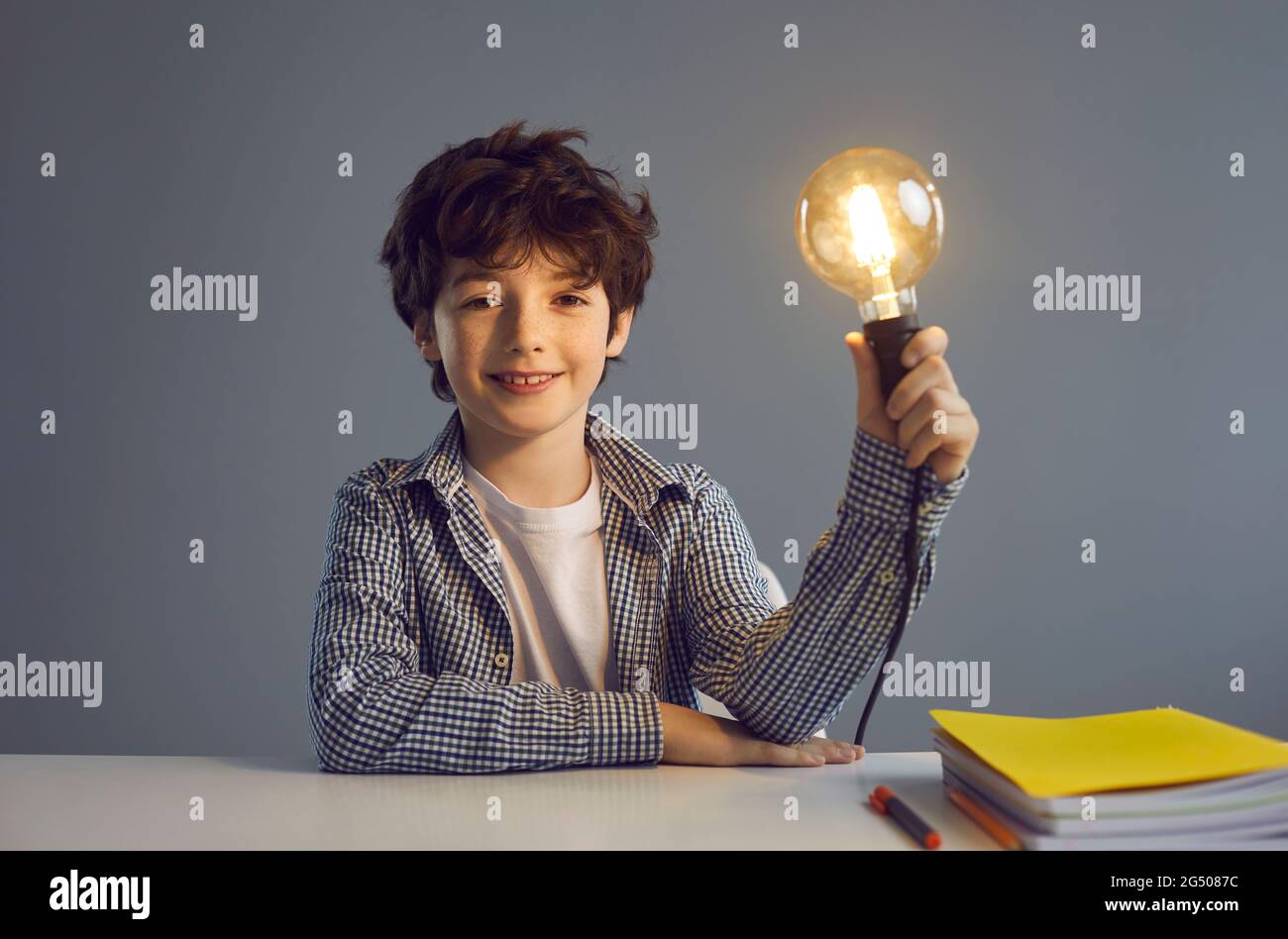 Smiling school boy child with light bulb sitting at desk against studio ...