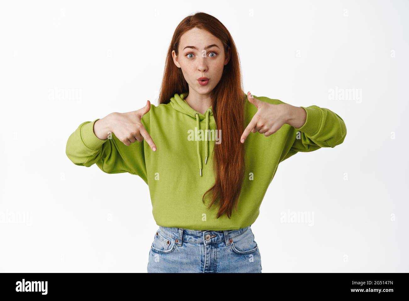 Surprised and curious redhead woman pointing fingers down, showing ...