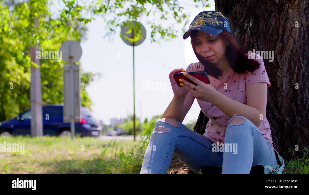 Young woman rests with mobile phone, sitting near tree on street ...